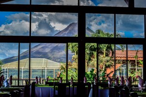 Restaurante com vista para montanhas e um estufa. Mesas dispostas com toalhas brancas e guardanapos roxos.