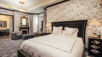 Um quarto espaçoso com uma cama grande, lareira, um espaço de estar com uma poltrona e uma toucador com espelho.