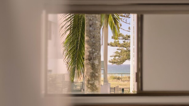 Quarto com uma janela grande, uma cama com roupa de cama branca e vista para a praia.