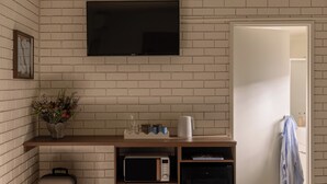 Quarto com parede de tijolos, televisão de tela plana, prateleira de madeira com um vaso de flores, micro-ondas, mala e porta com uma toalha pendurada.