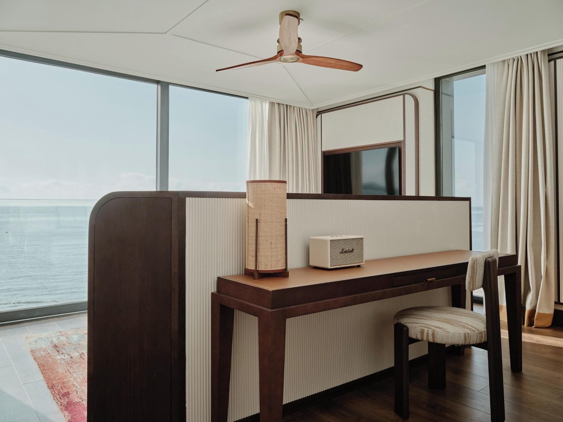 Quarto de hotel moderno com escrivaninha de madeira, uma cadeira pequena, um rádio e uma grande janela com vista para o mar.