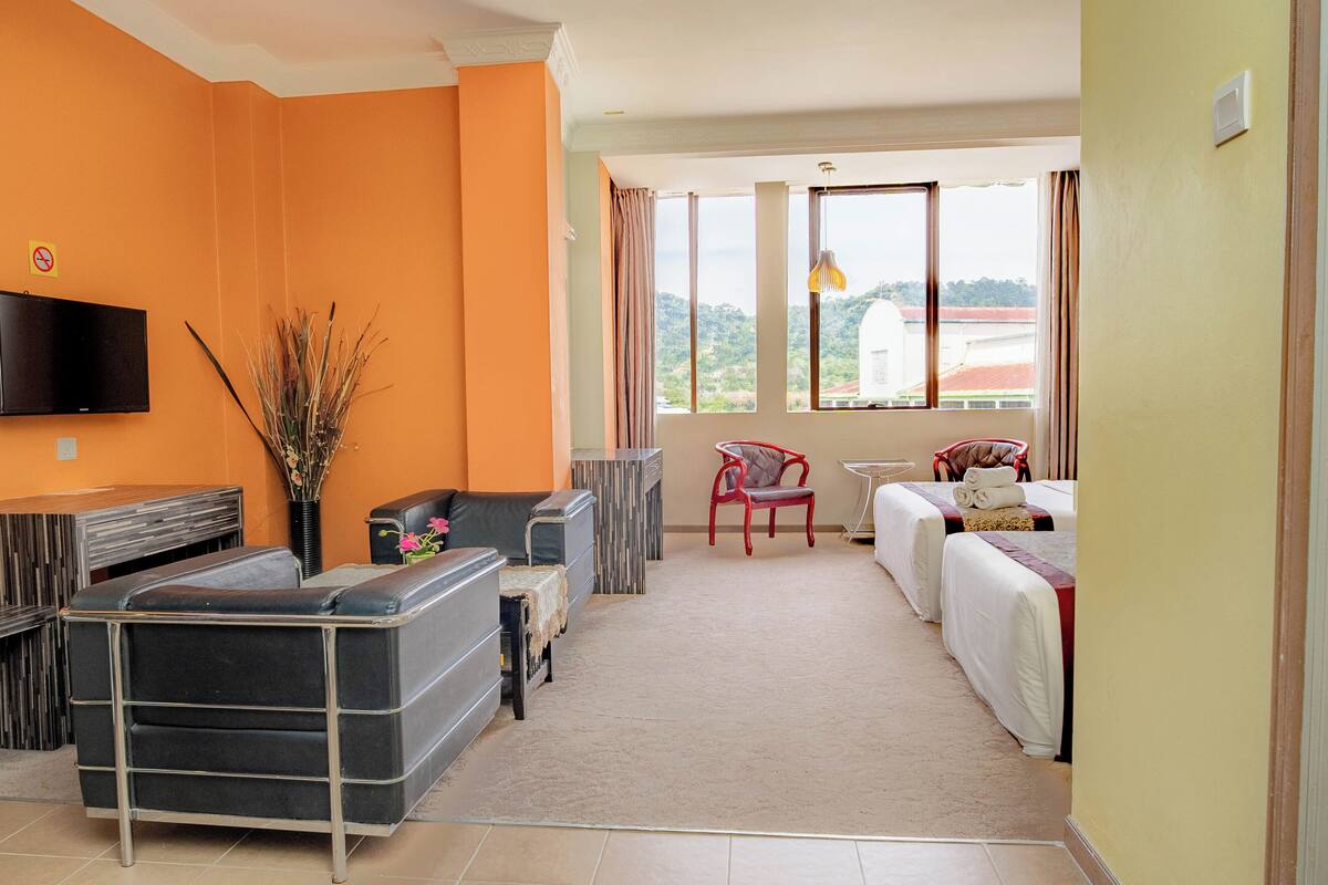 Quarto de hotel com sofá, poltrona, cama, televisão e vista para o exterior.