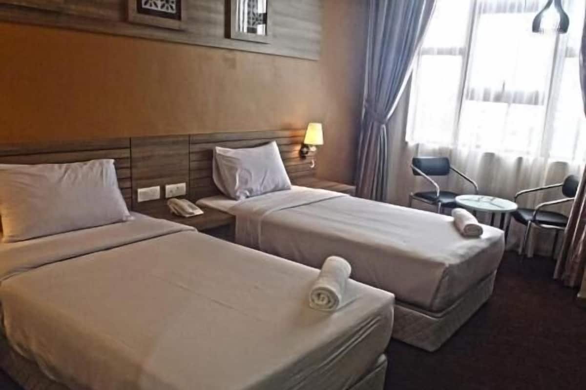 Quarto de hotel com duas camas, uma mesa de cabeceira com abajur, uma cadeira e uma pequena mesa com outra cadeira.