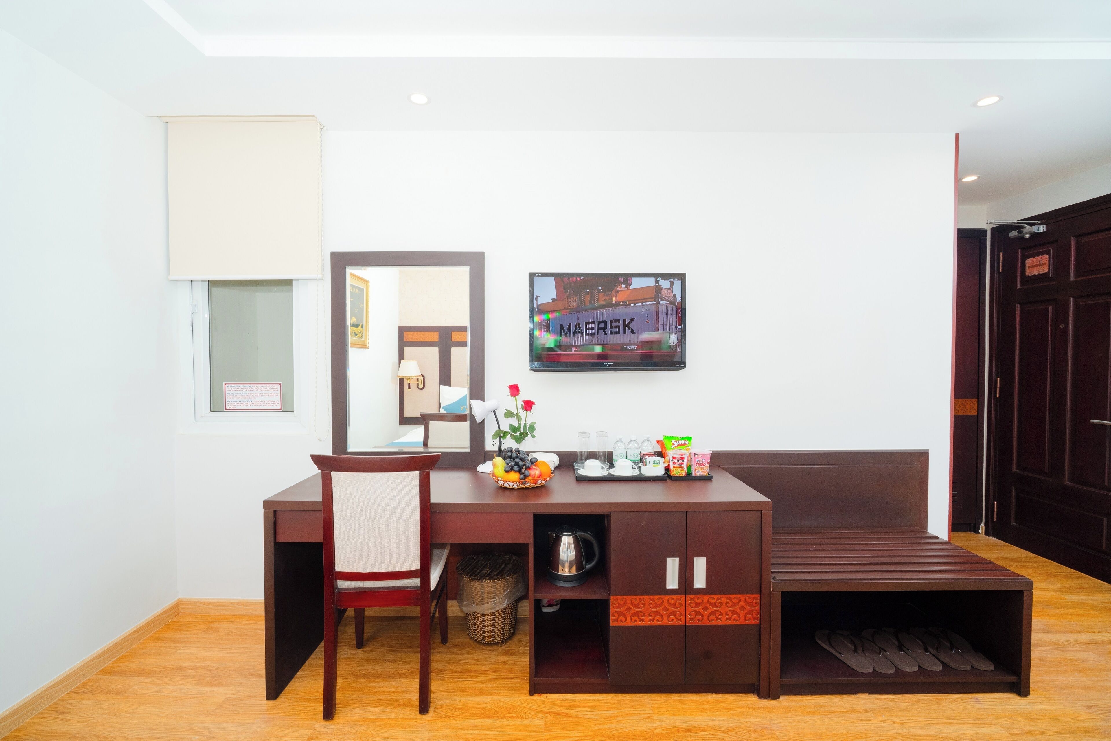 Quarto de hotel compacto com escrivaninha de madeira, uma cadeira, um pequeno sofá, televisão e espaço para guardar sapatos.