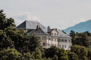 Um edifício grande e de vários andares com fachada simétrica, rodeado por vegetação densa e montanhas ao fundo.