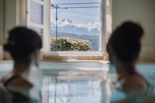 Duas pessoas em uma piscina com vista para montanhas nevadas através de uma janela.