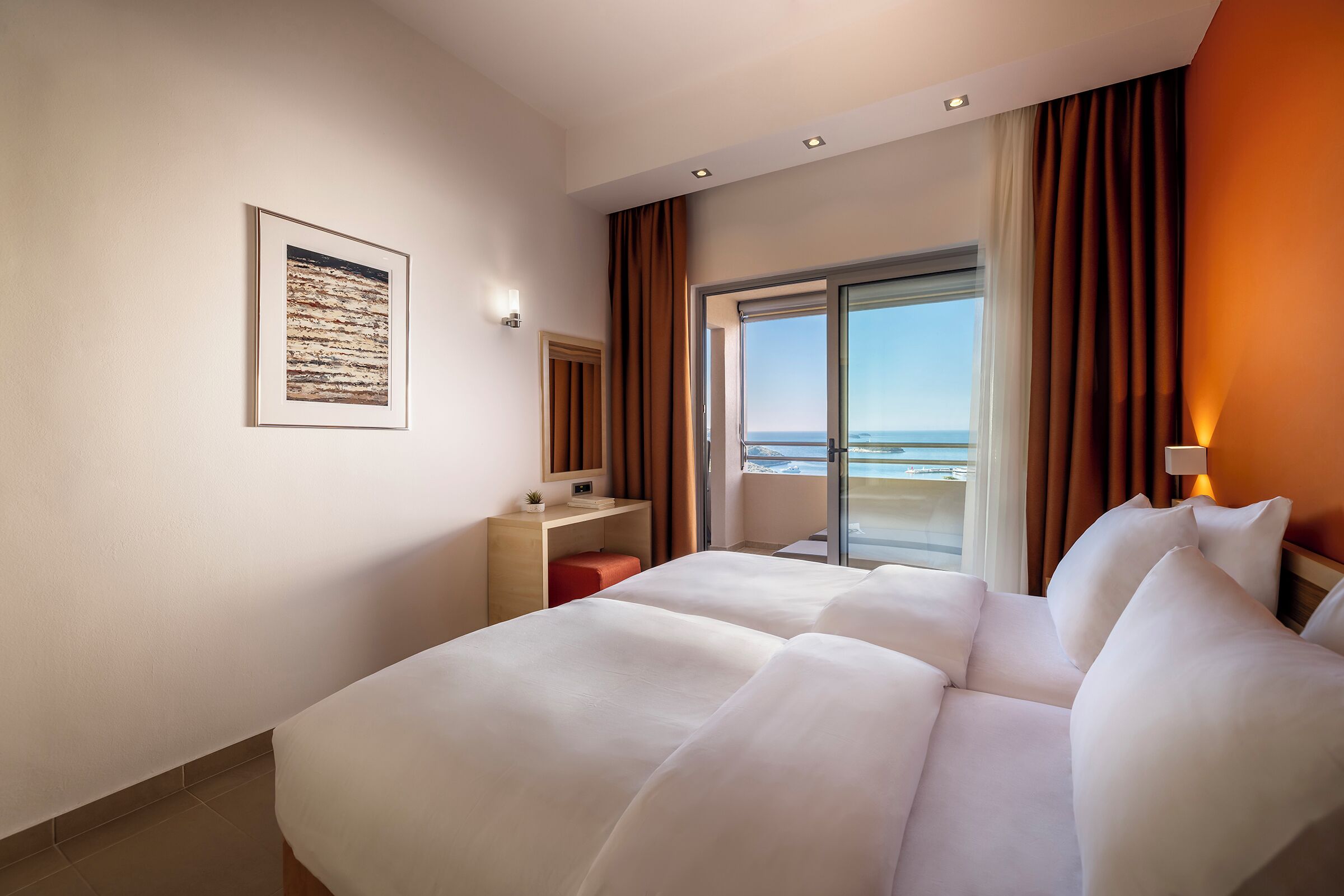 Quarto de hotel com uma cama grande, uma mesa pequena, uma janela vista para a praia e um quadro na parede.