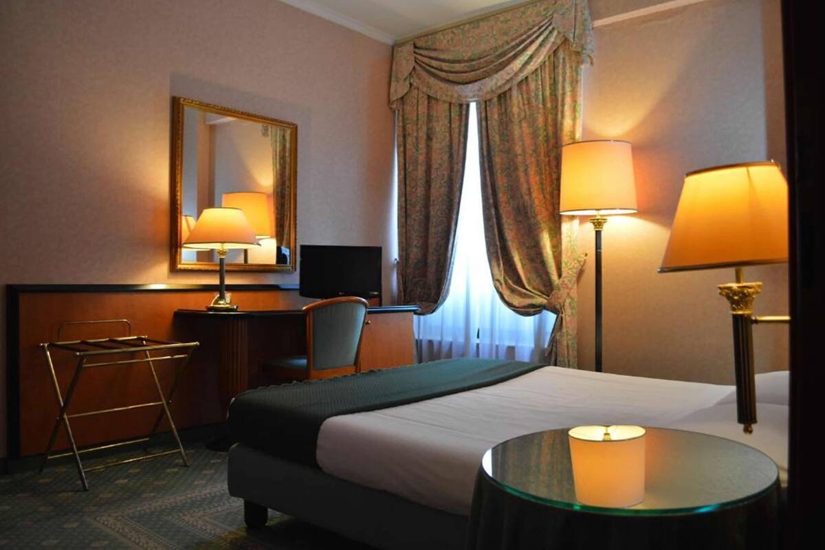 Quarto de hotel com cama, escrivaninha, cadeira, televisão e duas luminárias.