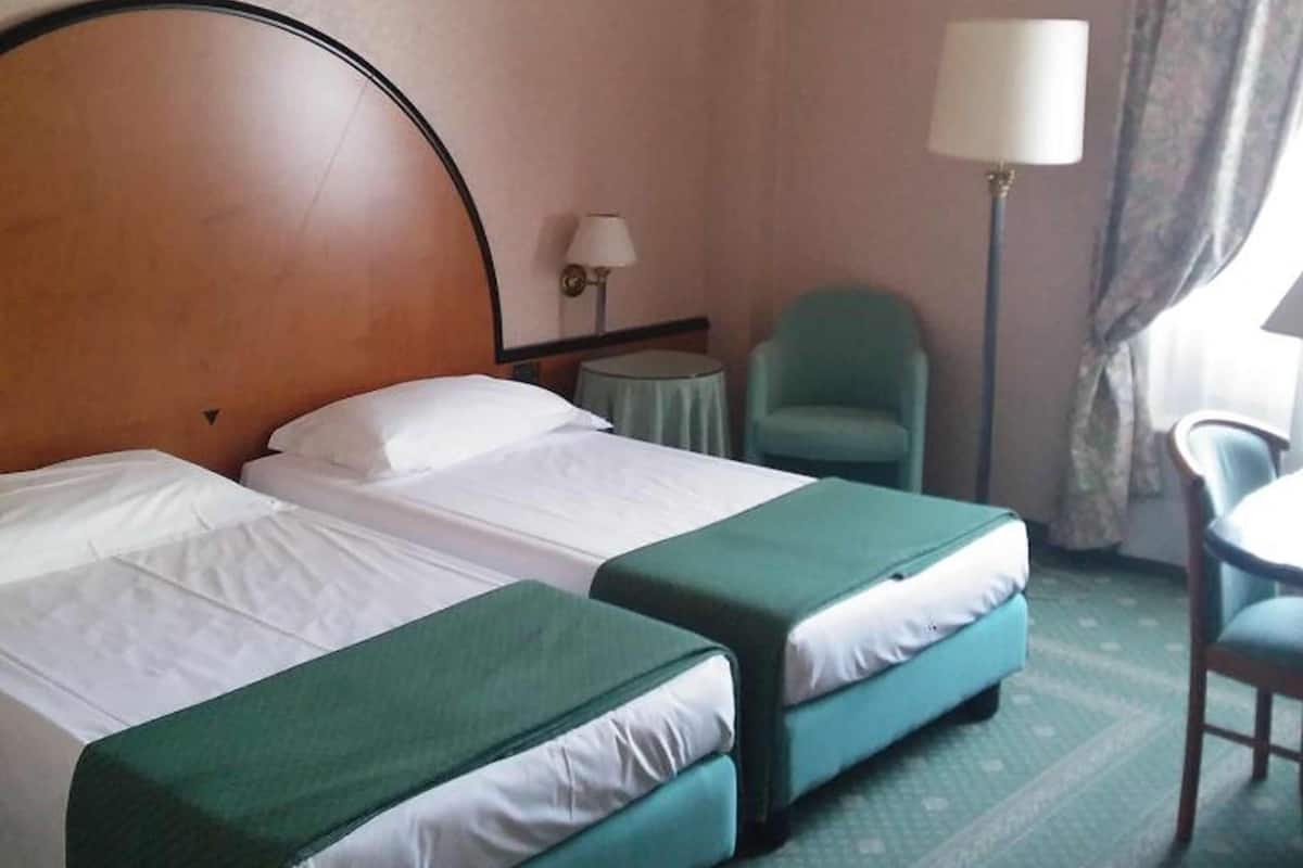 Quarto de hotel com duas camas, uma cadeira, um abajur e uma janela com cortinas.