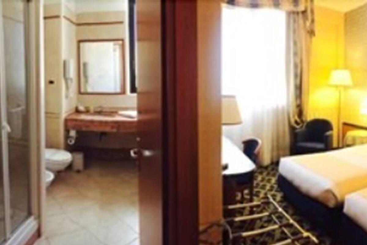 Quarto de hotel com cama, mesa de cabeceira, luminária e vista para o corredor.