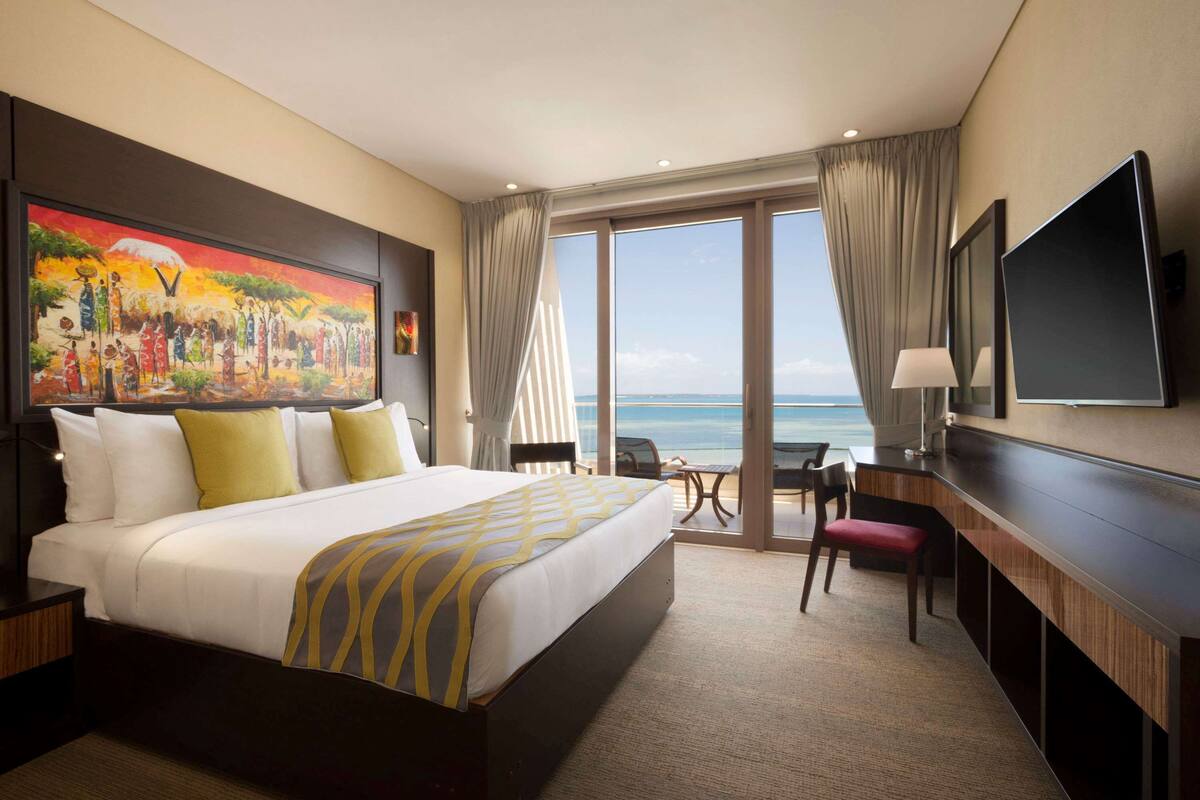 Quarto de hotel com cama grande, escrivaninha, cadeira, televisão e um quadro na parede.
