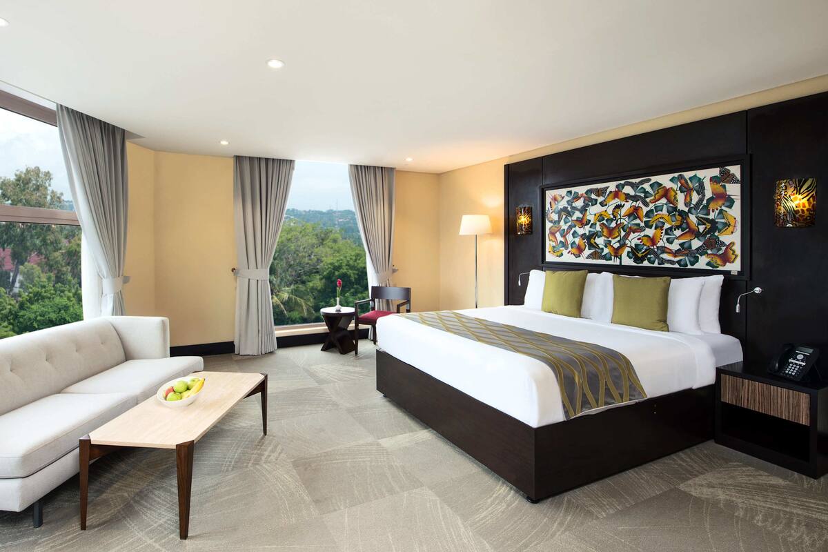 Quarto de hotel com cama, sofá, uma mesinha com frutas, vista para o exterior e um grande painel decorativo na parede.