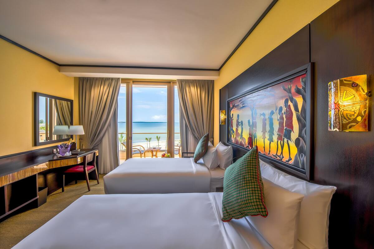 Quarto de hotel com duas camas, uma escrivaninha, uma cadeira e uma janela grande com vista para a praia.