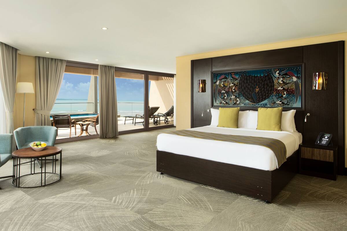 Quarto de hotel com uma cama grande, uma mesa, uma cadeira e vista para o oceano.