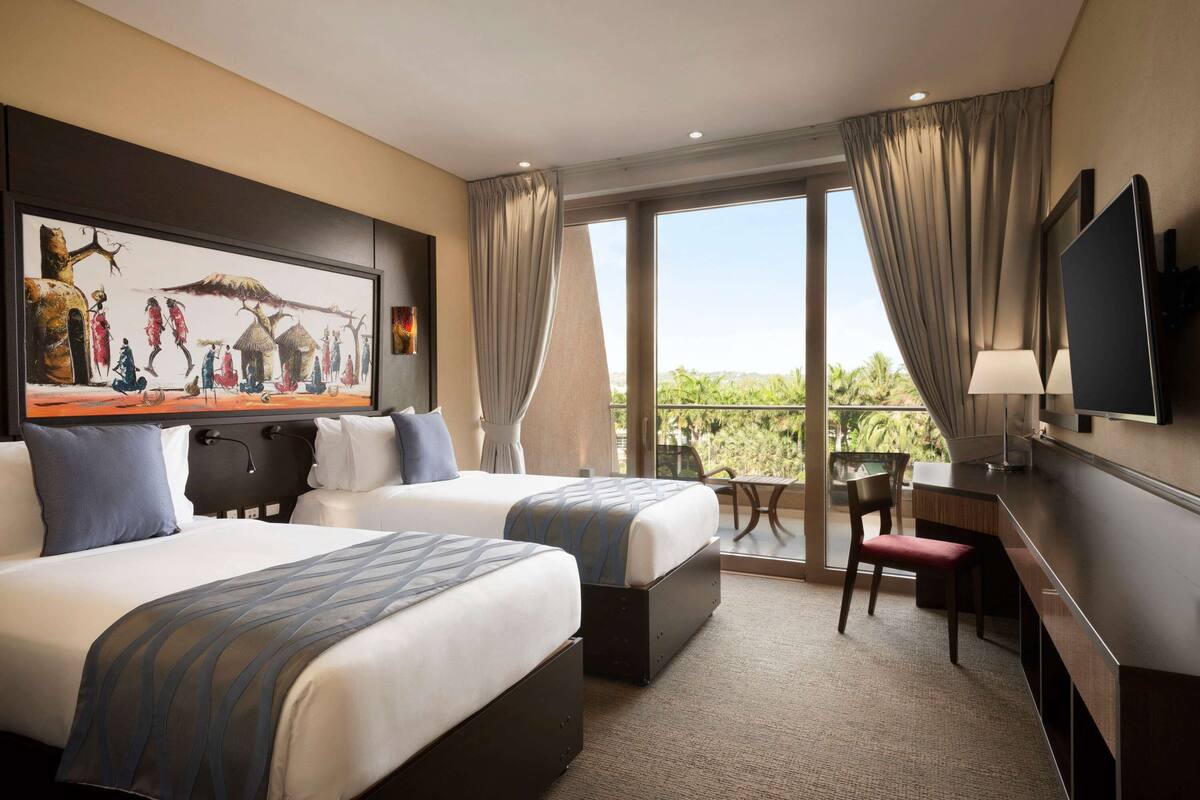 Quarto de hotel com duas camas, uma escrivaninha, uma cadeira, televisão e uma janela grande com cortinas.