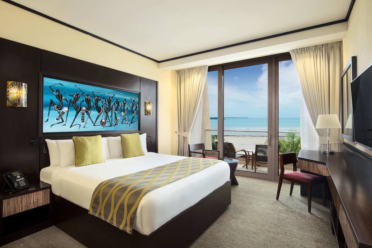 Quarto de hotel com uma cama grande, uma escrivaninha com cadeira, televisão e vista para o oceano através de uma porta de correr.