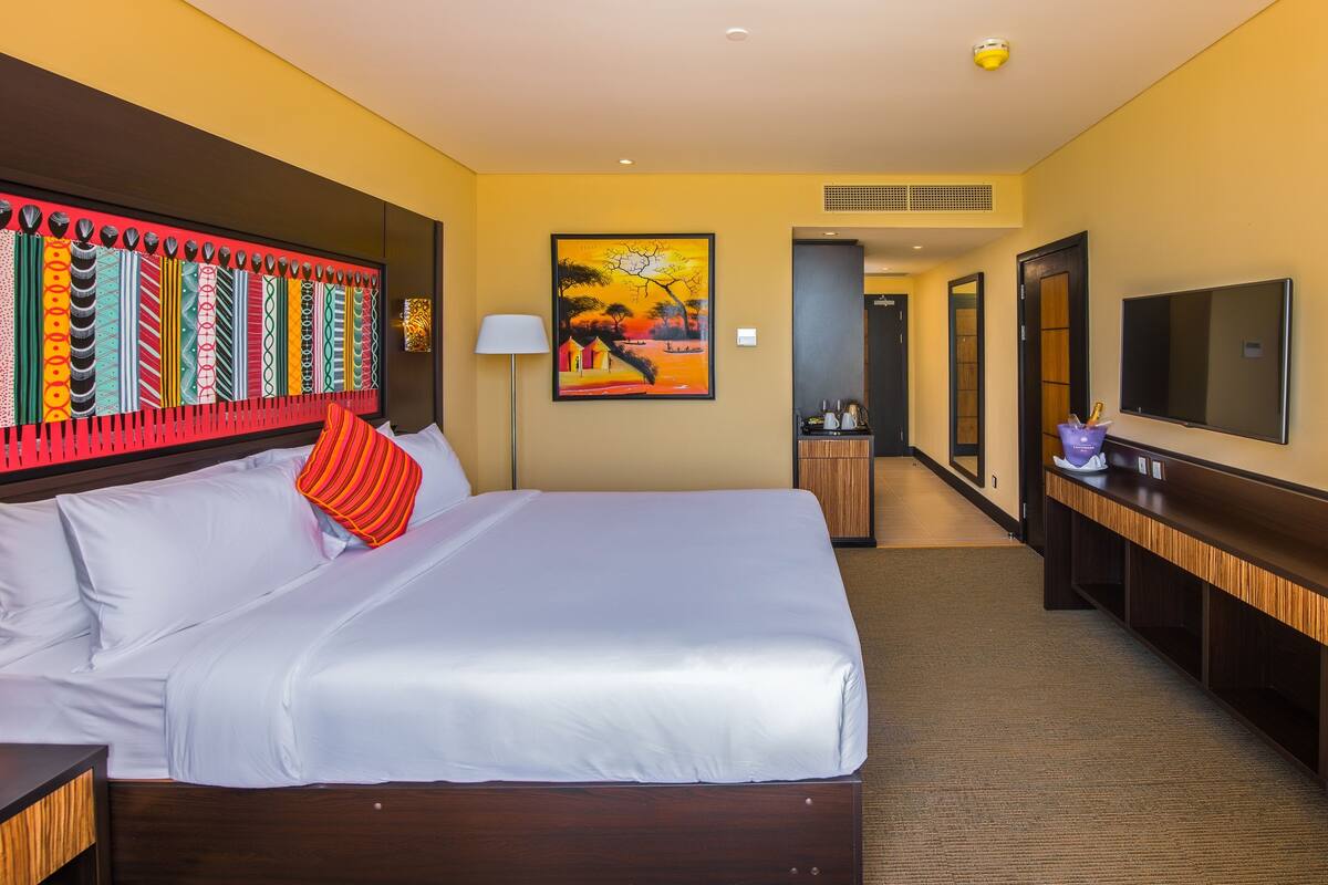 Quarto de hotel com uma cama grande, televisão presa à parede, escrivaninha com abajur e um quadro na parede.