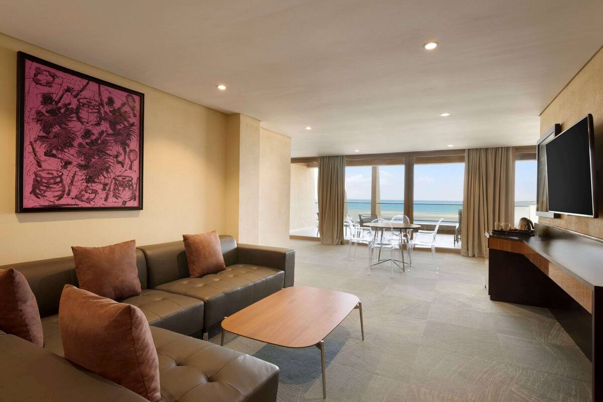 Quarto de hotel moderno com sofá, mesa de centro, TV e varanda com vista para o mar.