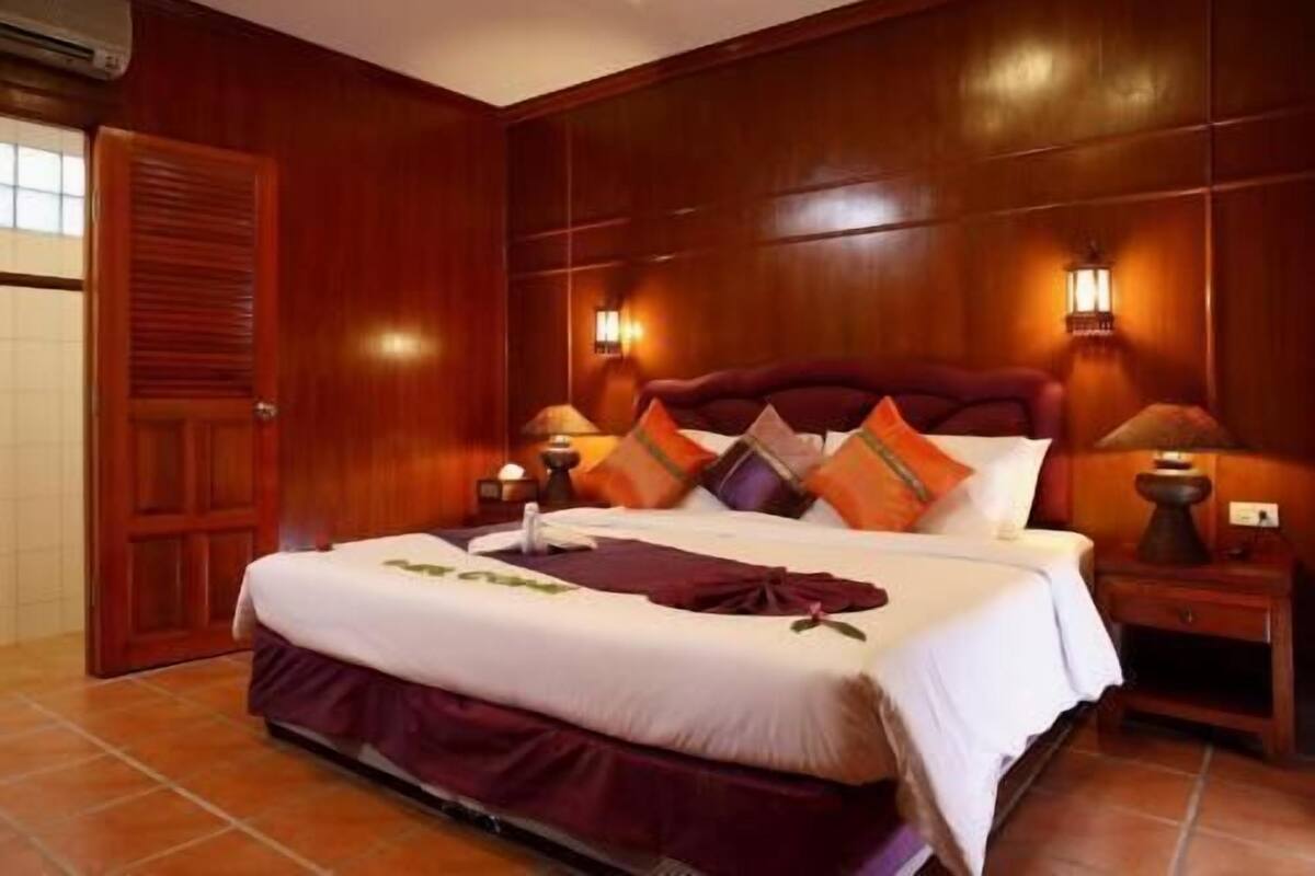 Quarto de hotel com cabeceira de madeira, cama com várias almofadas, duas luminárias de cabeceira e uma mesa de apoio.