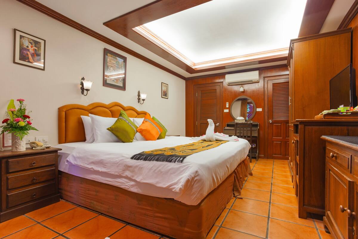 Quarto de hotel com uma cama grande, cabeceira de madeira e um claraboia. Há uma mesinha de cabeceira com uma luminária e um vaso de flores. O quarto possui móveis de madeira e piso de azulejo.