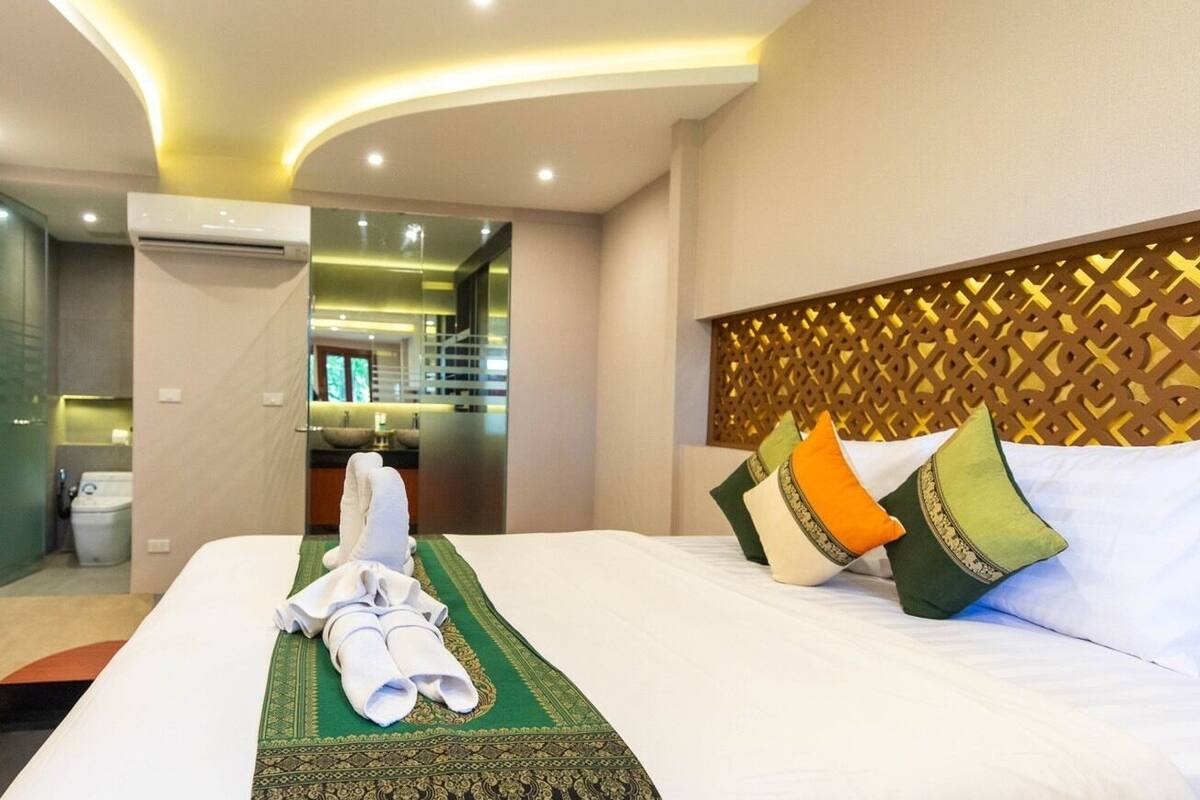 Quarto de hotel com cama bem arrumada, manta verde e preta com padrão e cabeceira de madeira.