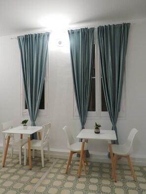 Quarto com duas mesas brancas e cadeiras, cada uma com uma pequena planta. O quarto tem um chão estampado e está decorado com cortinas azuis.