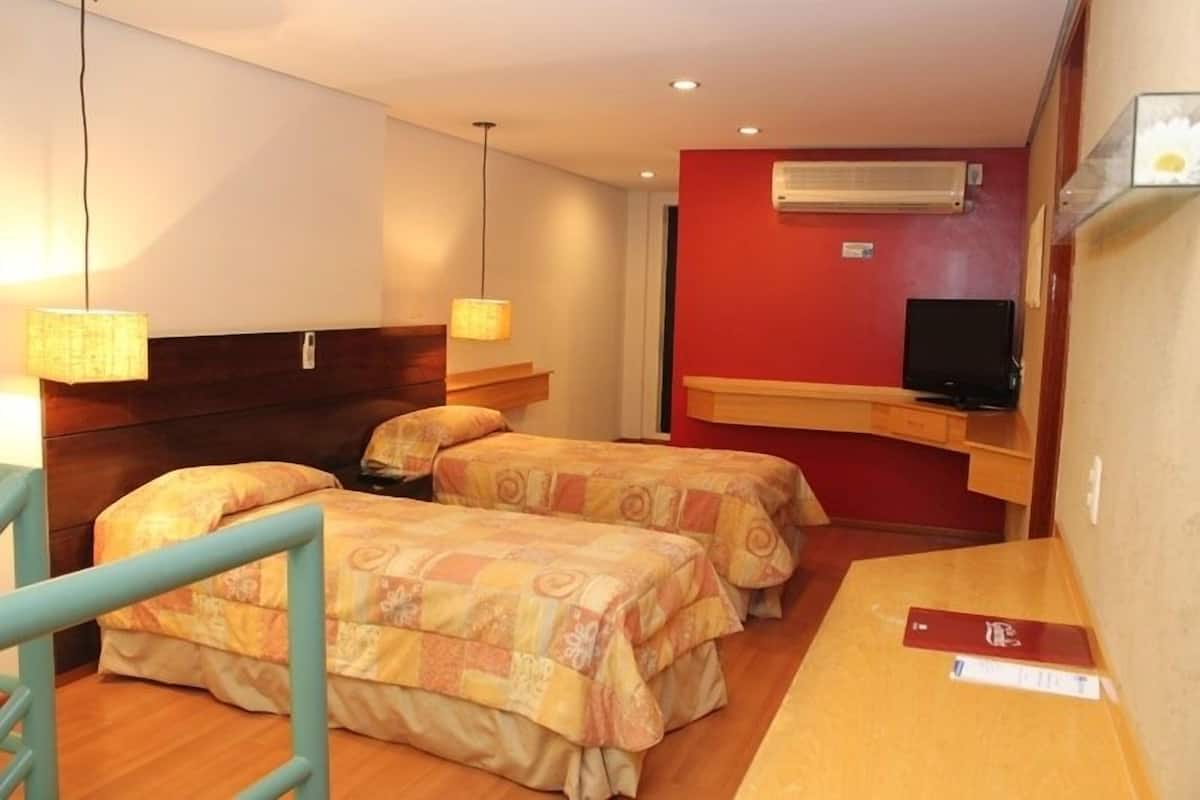 Quarto de hotel com duas camas, uma escrivaninha, uma televisão e aparelho de ar condicionado.