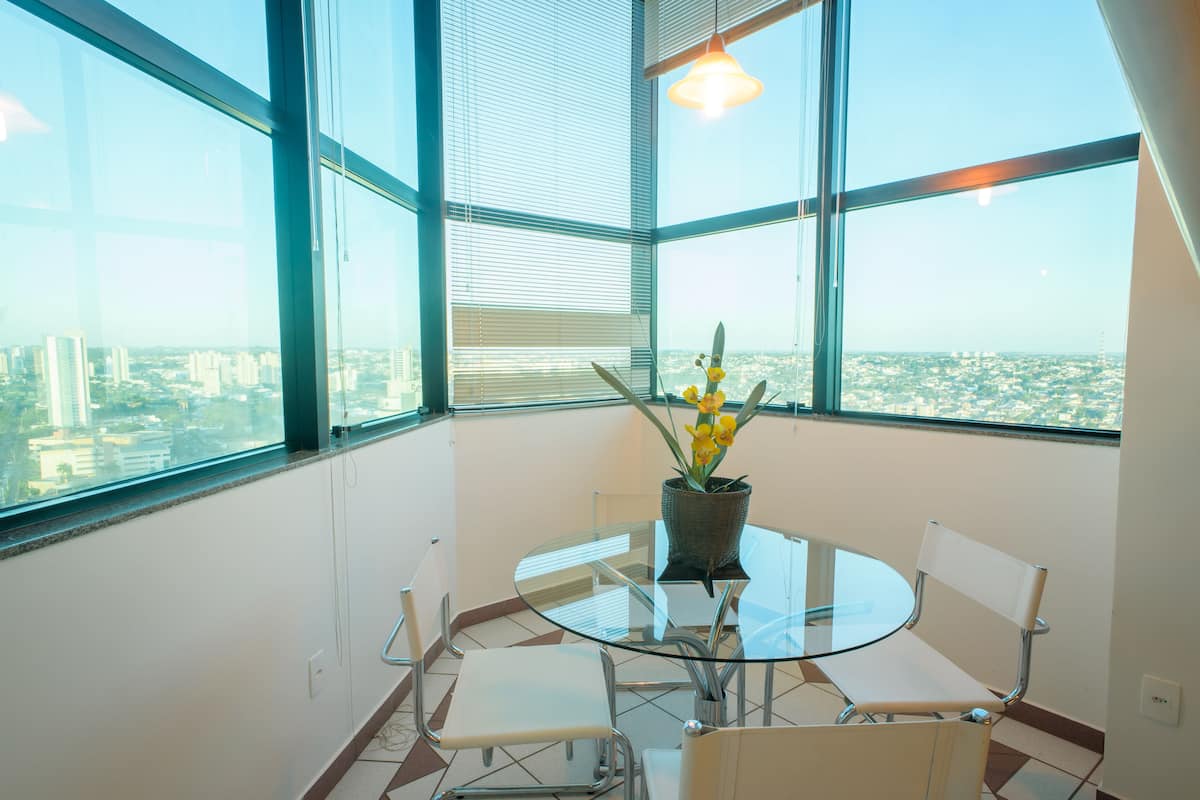 Quarto moderno com mesa e cadeiras de vidro, uma planta em vaso e janelas amplas com vista para a cidade.