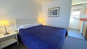 Quarto de hotel com cama, duas luminárias laterais, um quadro na parede e vista para a área da cozinha.