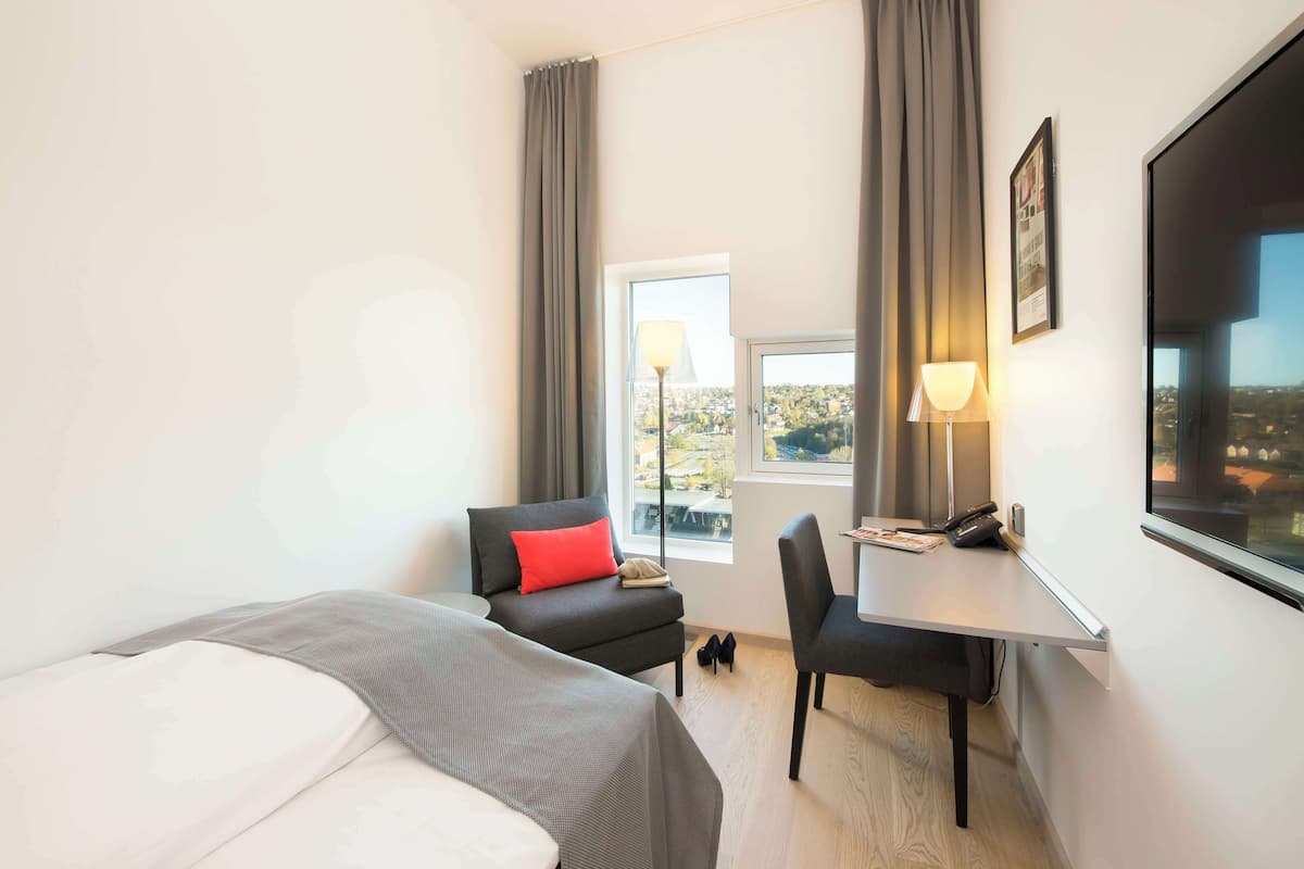 Quarto de hotel com cama, escrivaninha, cadeira, janela com vista e luminária.