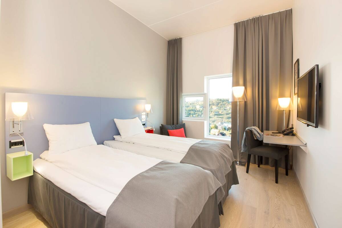 Quarto de hotel com duas camas, uma escrivaninha, uma cadeira, uma televisão e uma janela com cortinas.