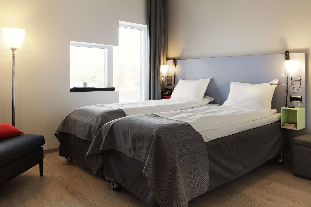 Quarto de hotel com duas camas, uma escrivaninha, uma cadeira e uma janela com cortinas.