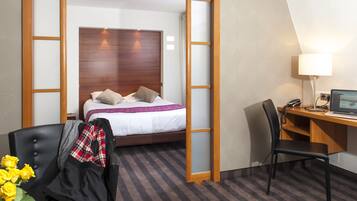 Quarto de hotel com uma cama, uma escrivaninha com computador, uma cadeira e uma luminária.