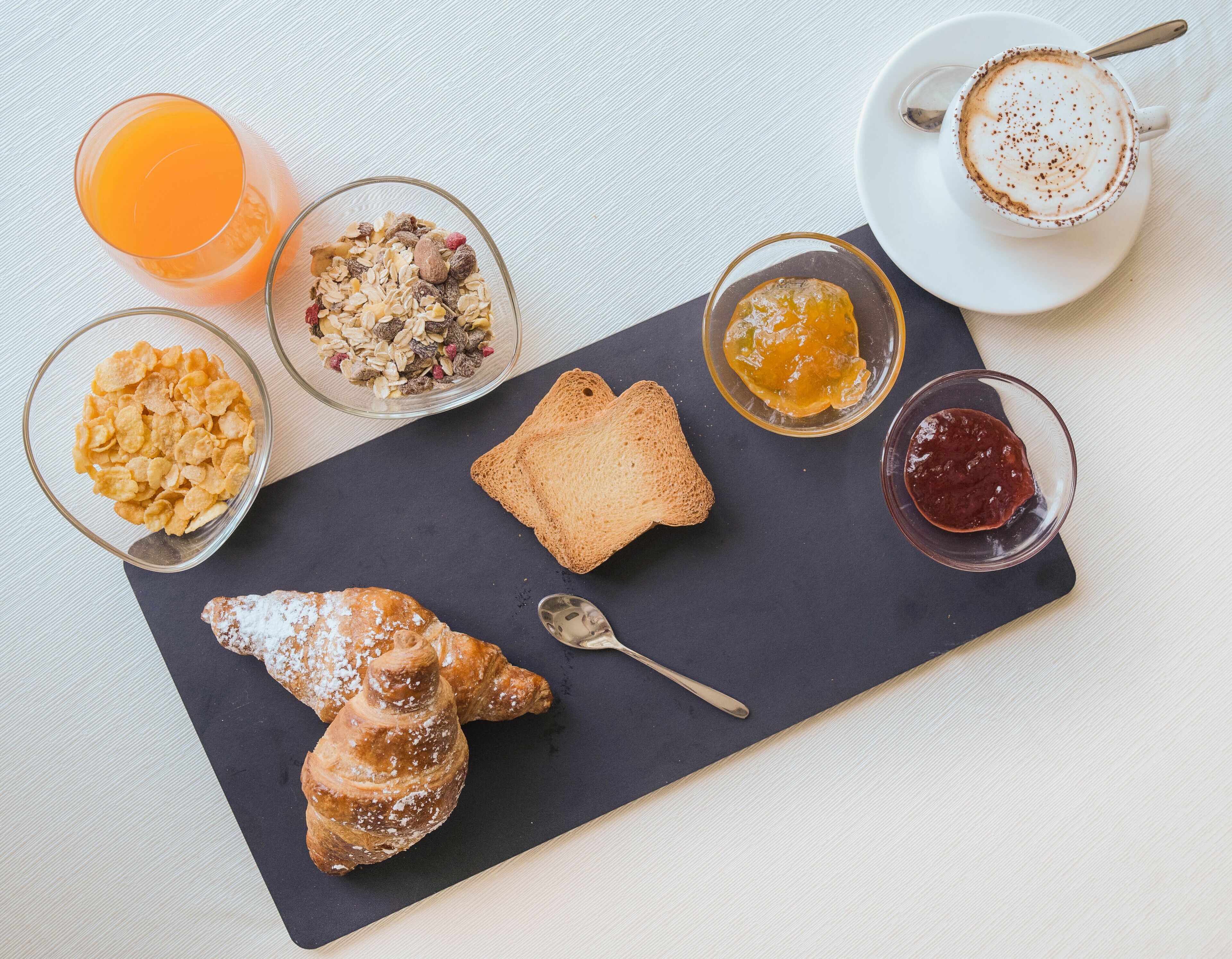 Disposição de café da manhã com croissant, torradas, cereal, geleia e café em uma lousa.
