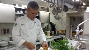 Um chef em cozinha profissional, com eletrodomésticos de aço inoxidável, diversos utensílios de cozinha e uma planta de ervas em vaso.