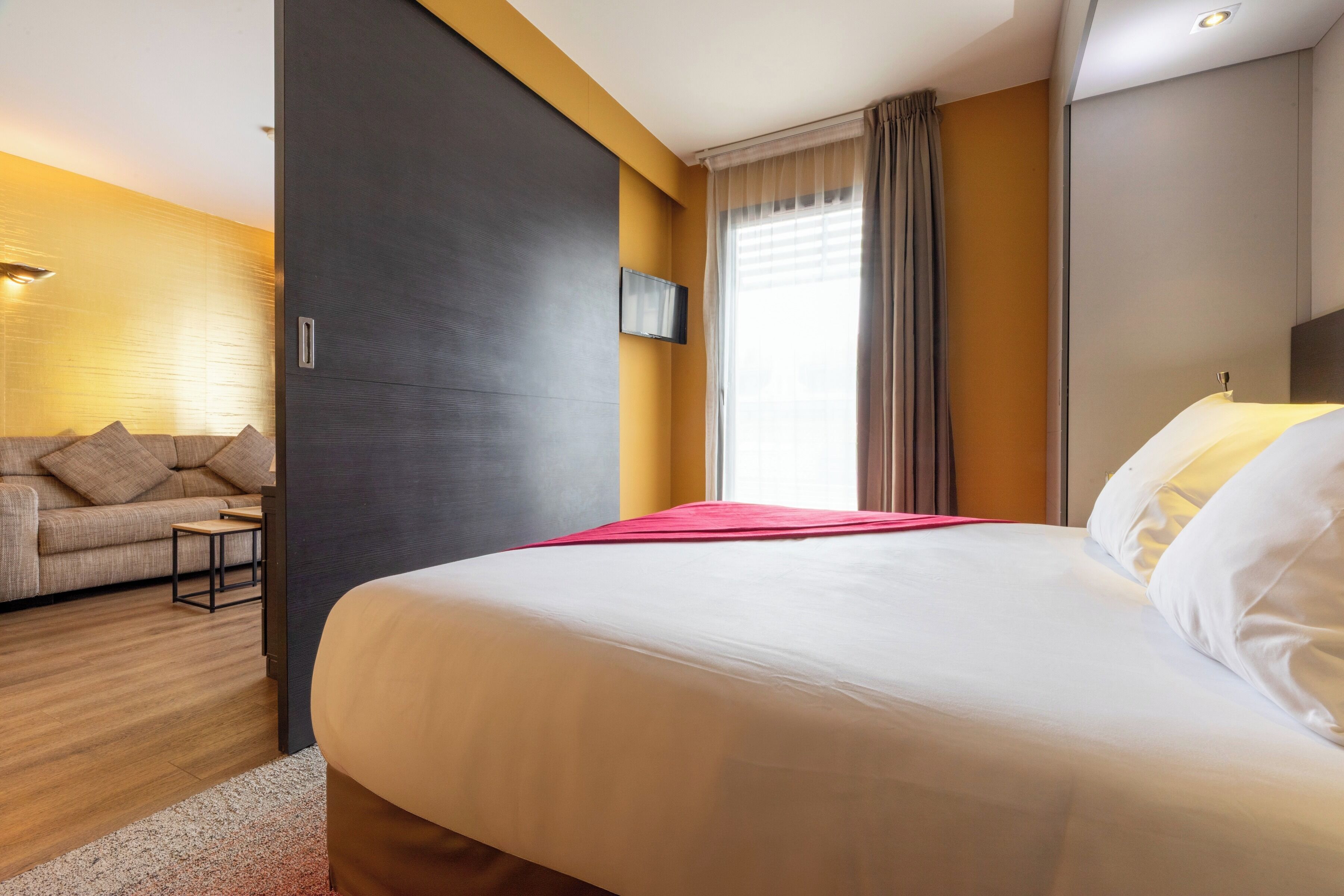 Quarto de hotel com cama, sofá, televisão e janela com cortinas.