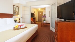 Quarto de hotel com cama, televisão, escrivaninha e banheiro visível através de uma porta aberta.