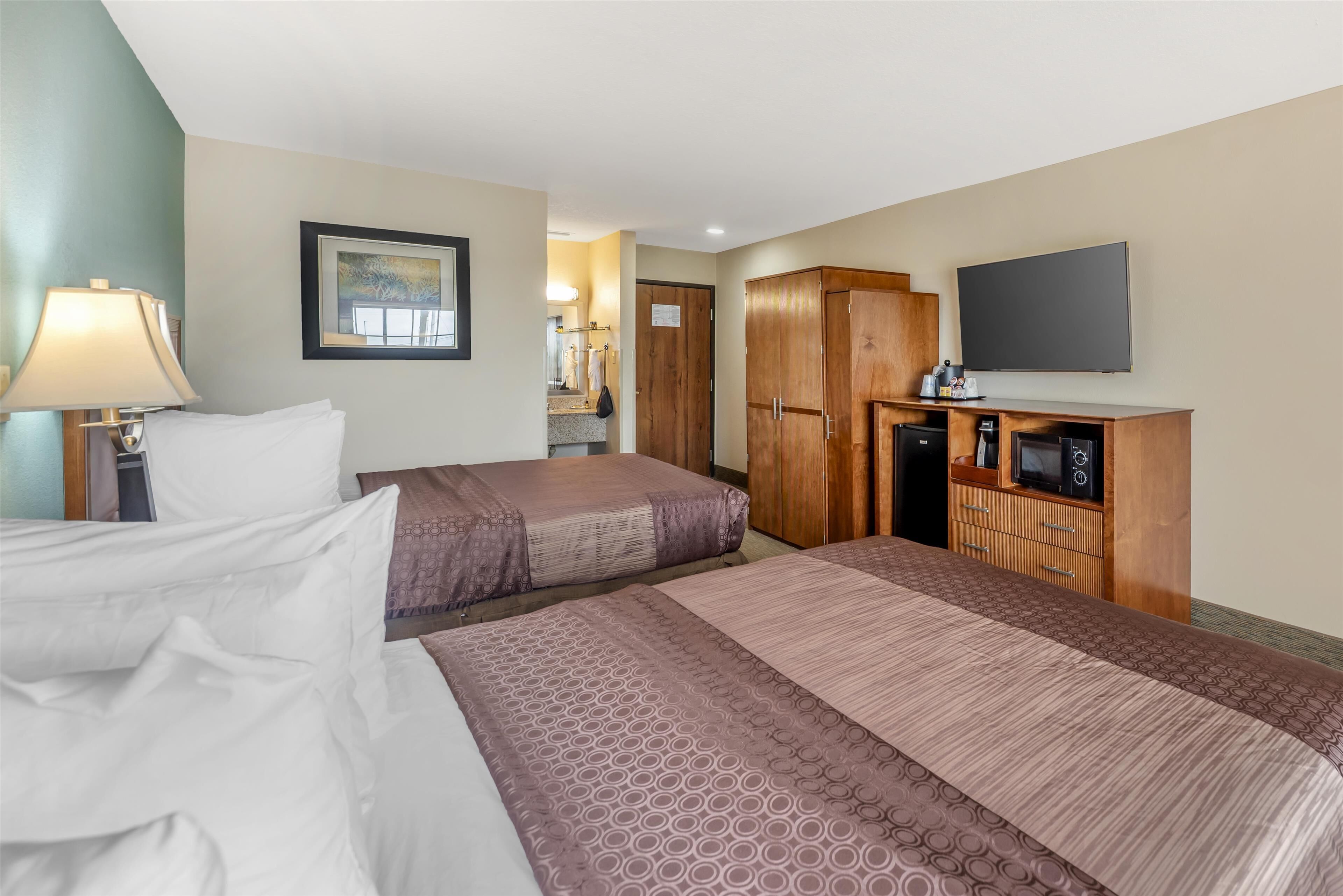 Quarto de hotel com duas camas, televisão, secretária com micro-ondas e casa de banho visível ao fundo.