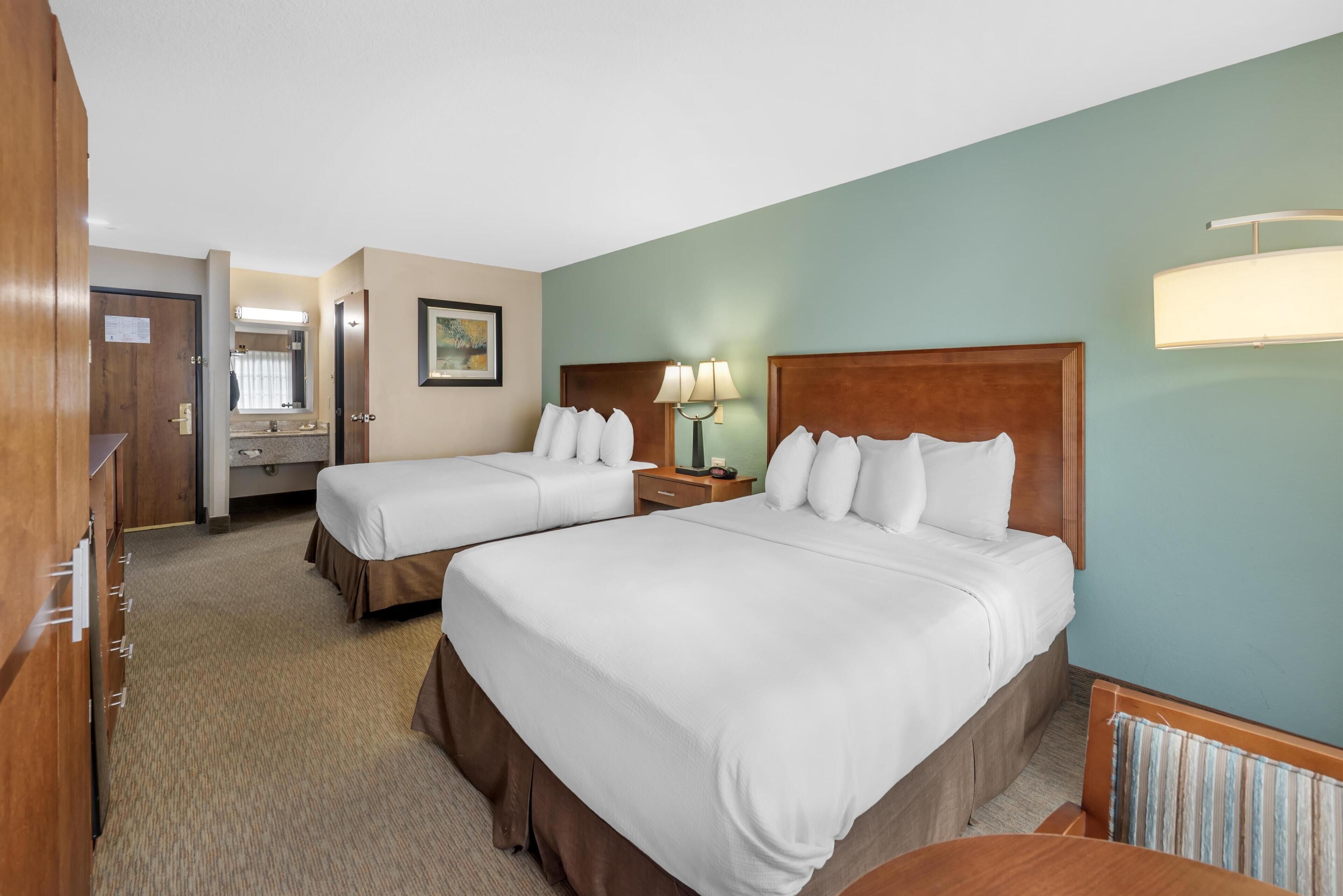 Quarto de hotel com duas camas, uma mesa, uma cadeira, uma luminária e um banheiro visível através de uma porta aberta.