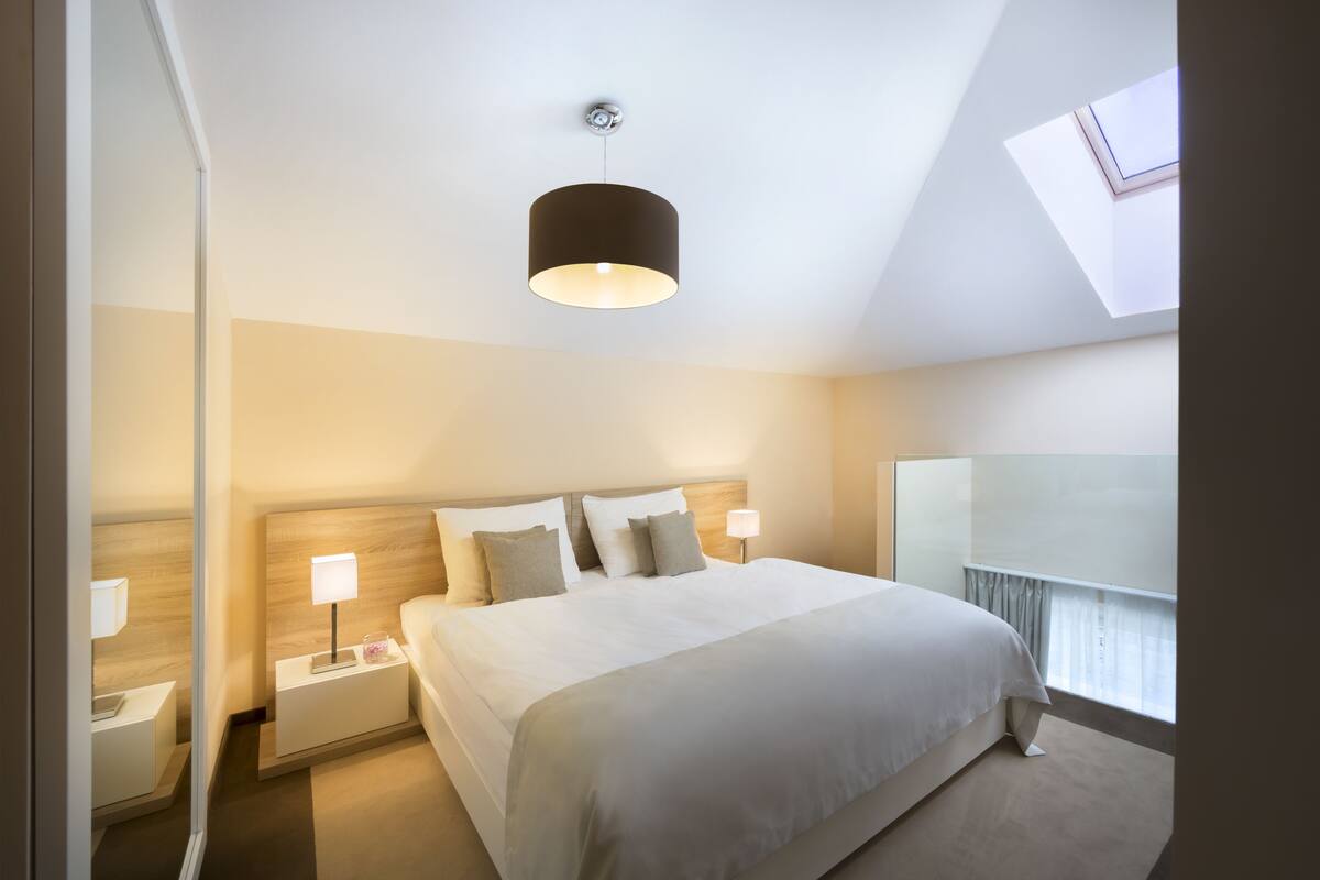 Quarto de hotel moderno com uma cama grande, mesa de cabeceira com abajur, iluminação no teto e uma janela ampla com cortinas.