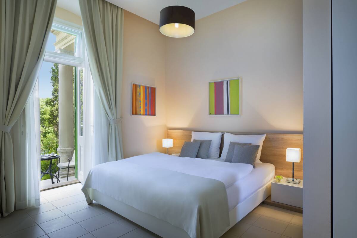 Quarto de hotel com uma cama grande, duas mesas de cabeceira com abajures, uma janela com cortinas e duas pinturas abstratas na parede.