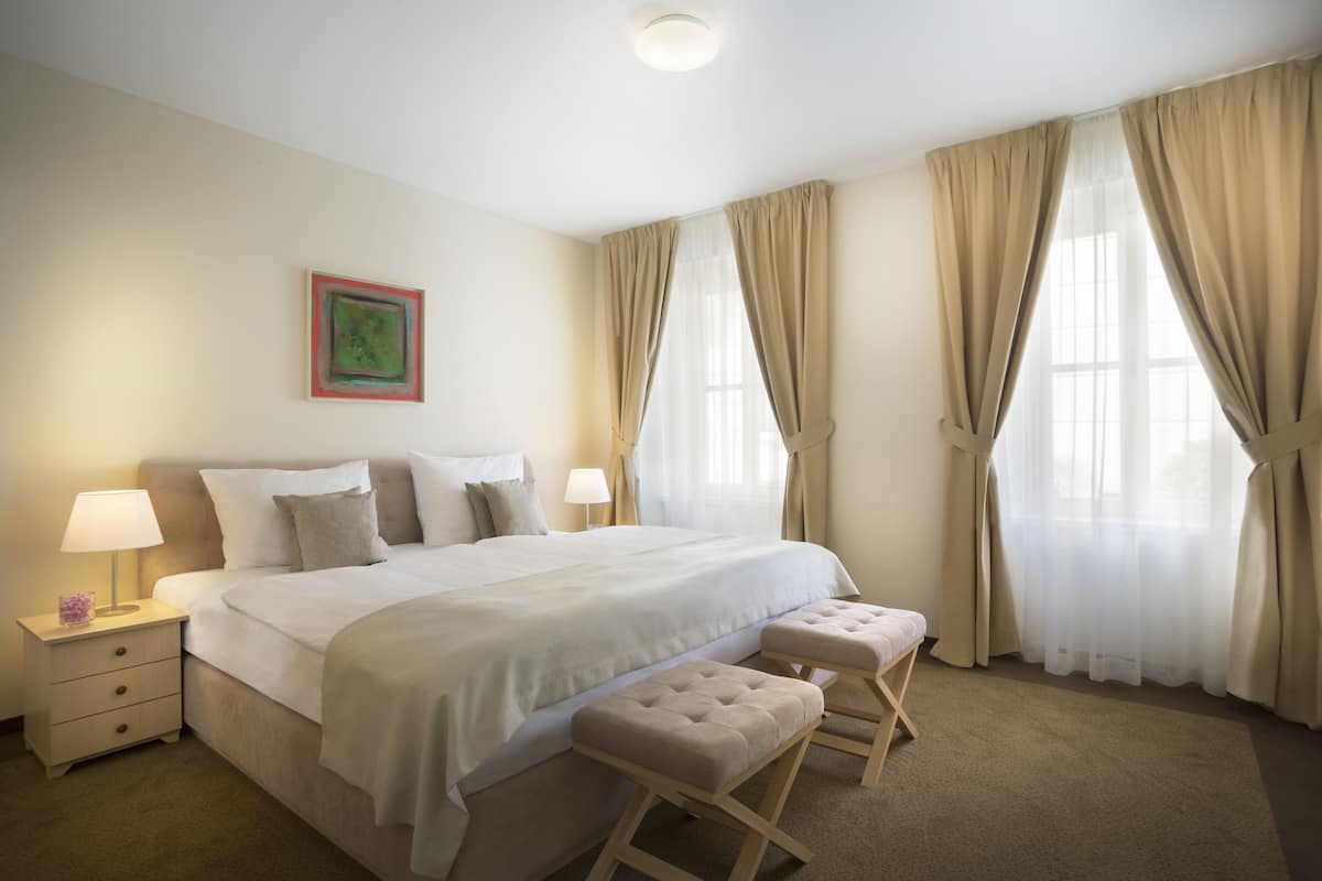 Quarto de hotel com uma cama grande, duas mesas de cabeceira com abajures, um banco e uma janela com cortinas.