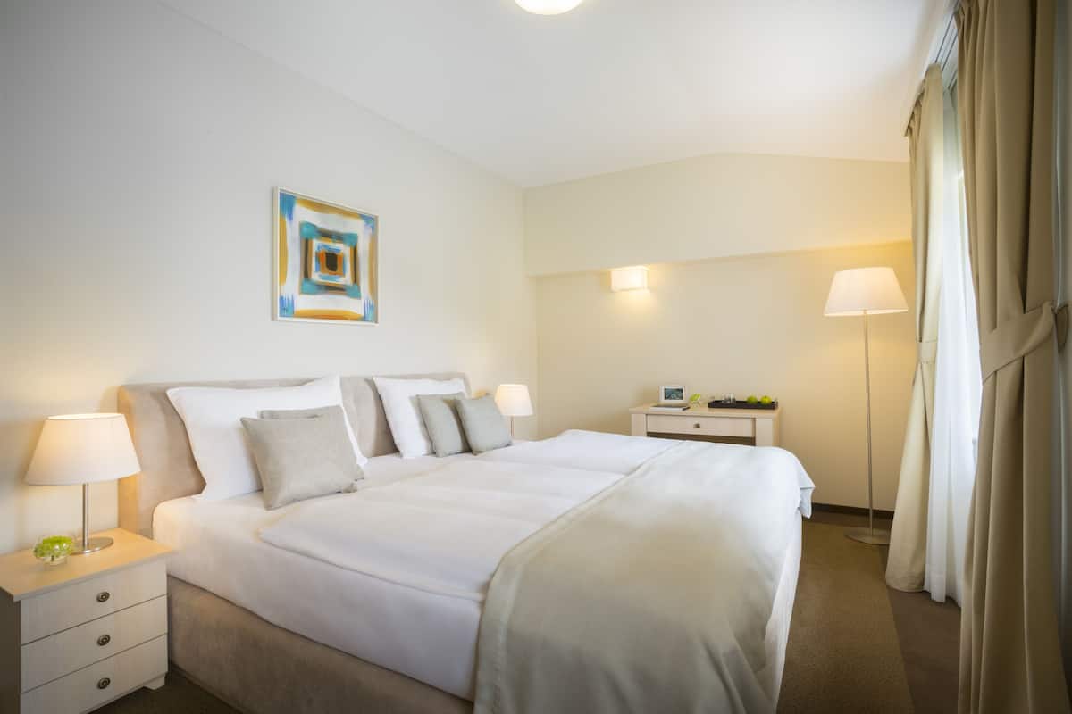 Quarto de hotel com uma cama grande, duas mesas de cabeceira, um criado-mudo, uma luminária na parede e um abajur, além de um quadro na parede.