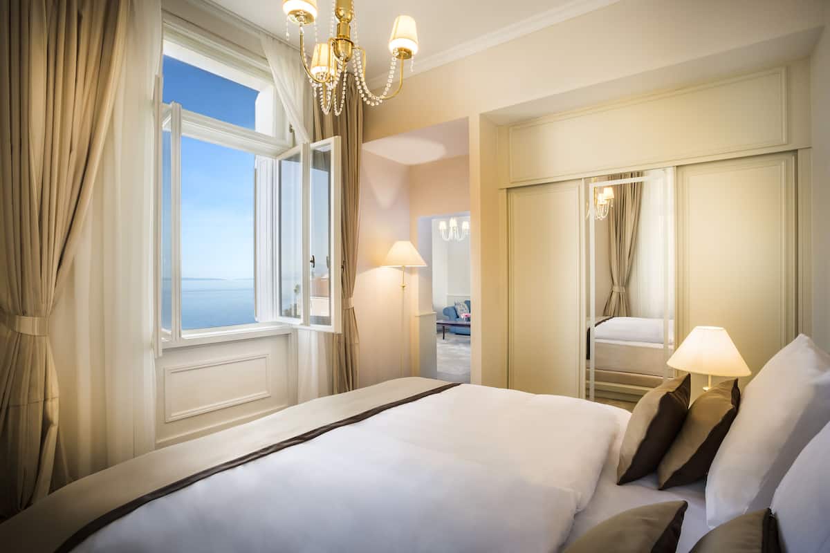 Quarto de hotel com uma cama grande, um candelabro, vista pela janela aberta e um banheiro visível ao fundo.