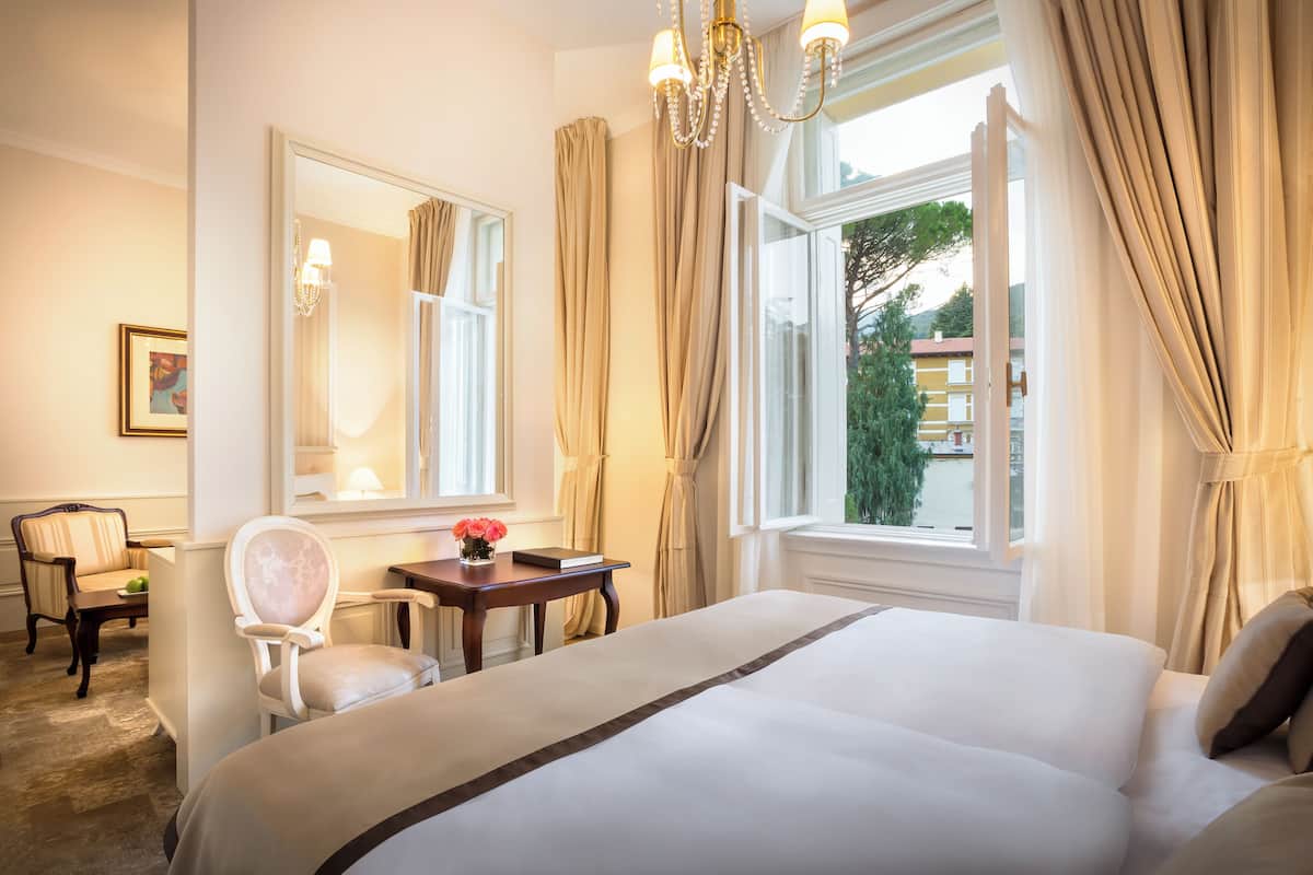 Quarto de hotel com uma cama grande, uma escrivaninha com cadeira, um espelho e uma janela com cortinas.