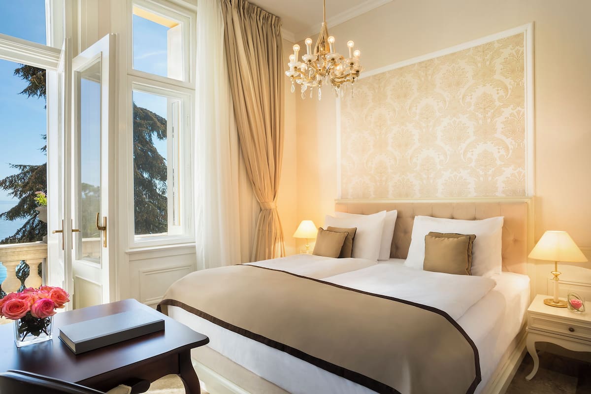 Quarto de hotel com uma cama grande, duas mesas de cabeceira, um candelabro e vista para o exterior.