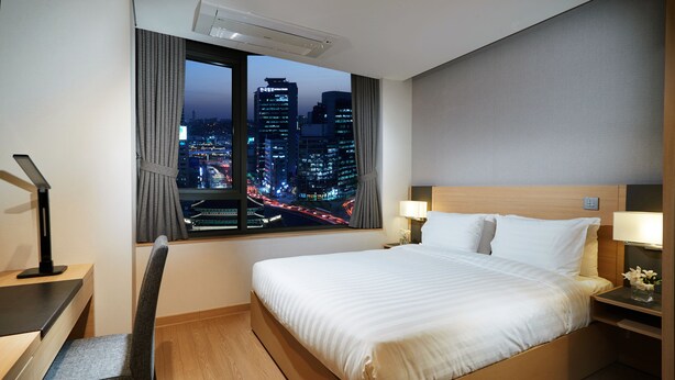 Quarto de hotel com cama, escrivaninha, cadeira e vista para o horizonte noturno.