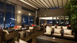 Um lounge no terraço com móveis de vime e amplas janelas com vista para a cidade.