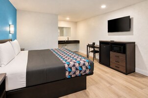 Quarto de hotel com cama, escrivaninha, cadeira, televisão e cozinha compacta.