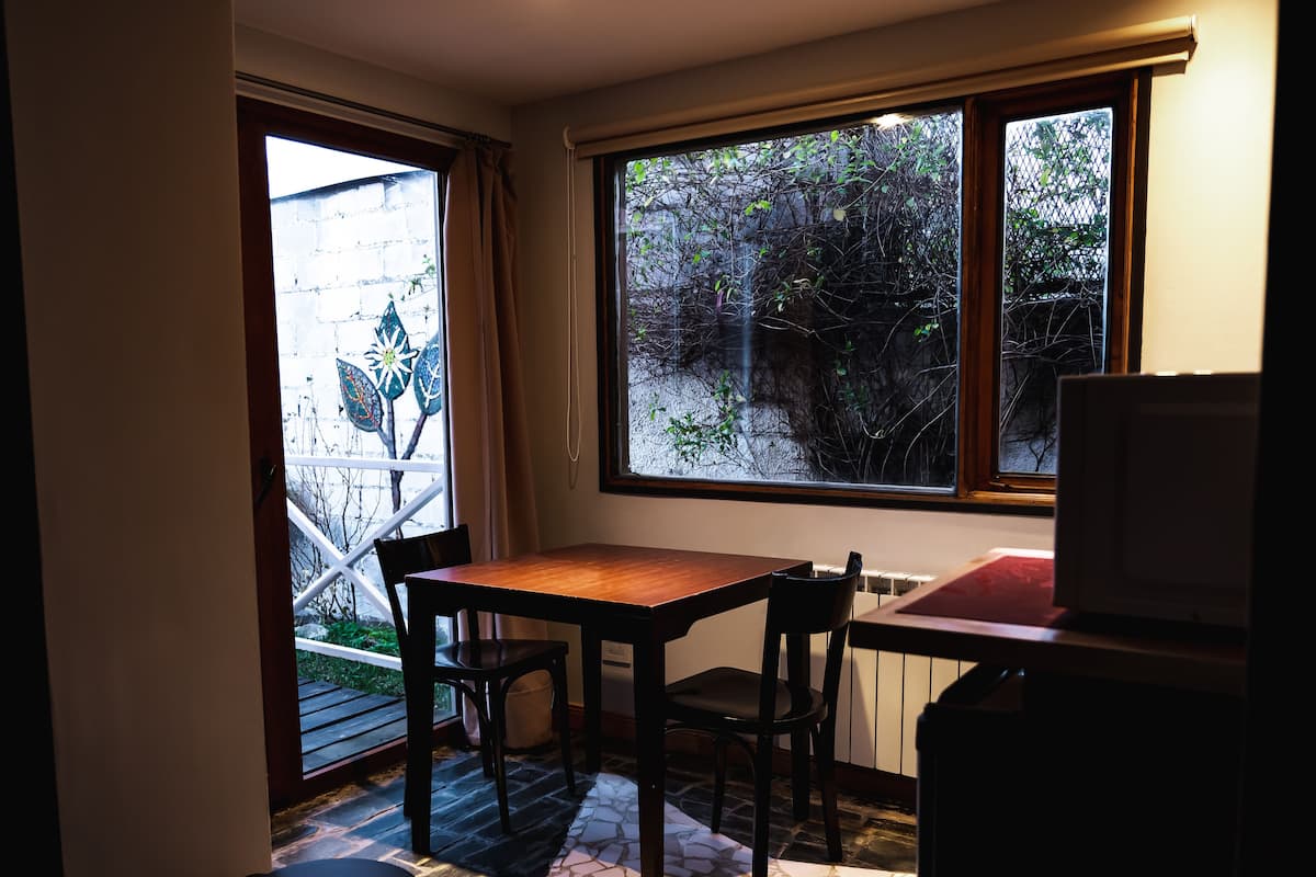 Uma pequena área de refeições com mesa e cadeiras, uma janela com vista para um jardim e um radiador sob a janela.